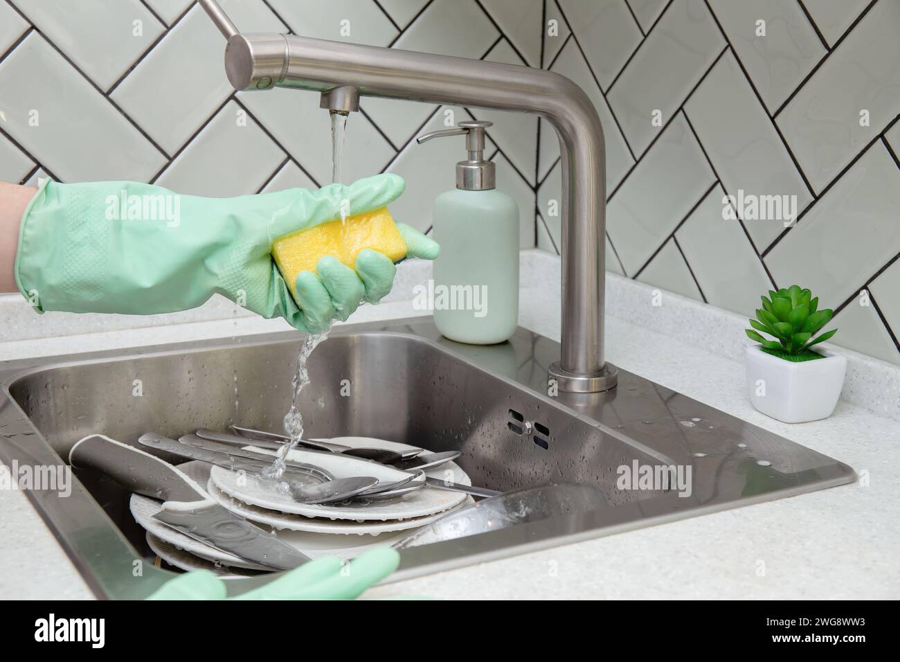 Hand in green glove squeezes sponge under faucet. Plates and cutlery ...