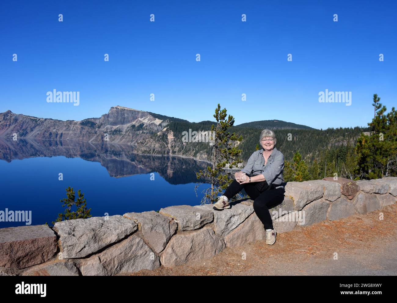 Happy visitor sits on rock wall smiling. Behind her is the view she is ...