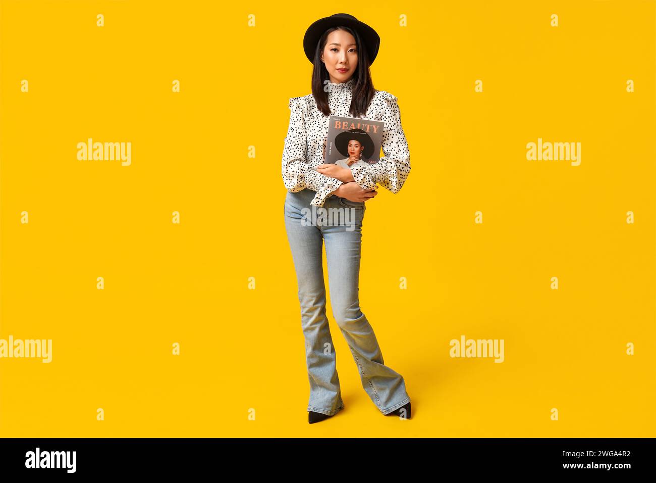Beautiful young Asian woman in stylish jeans with magazine on yellow ...