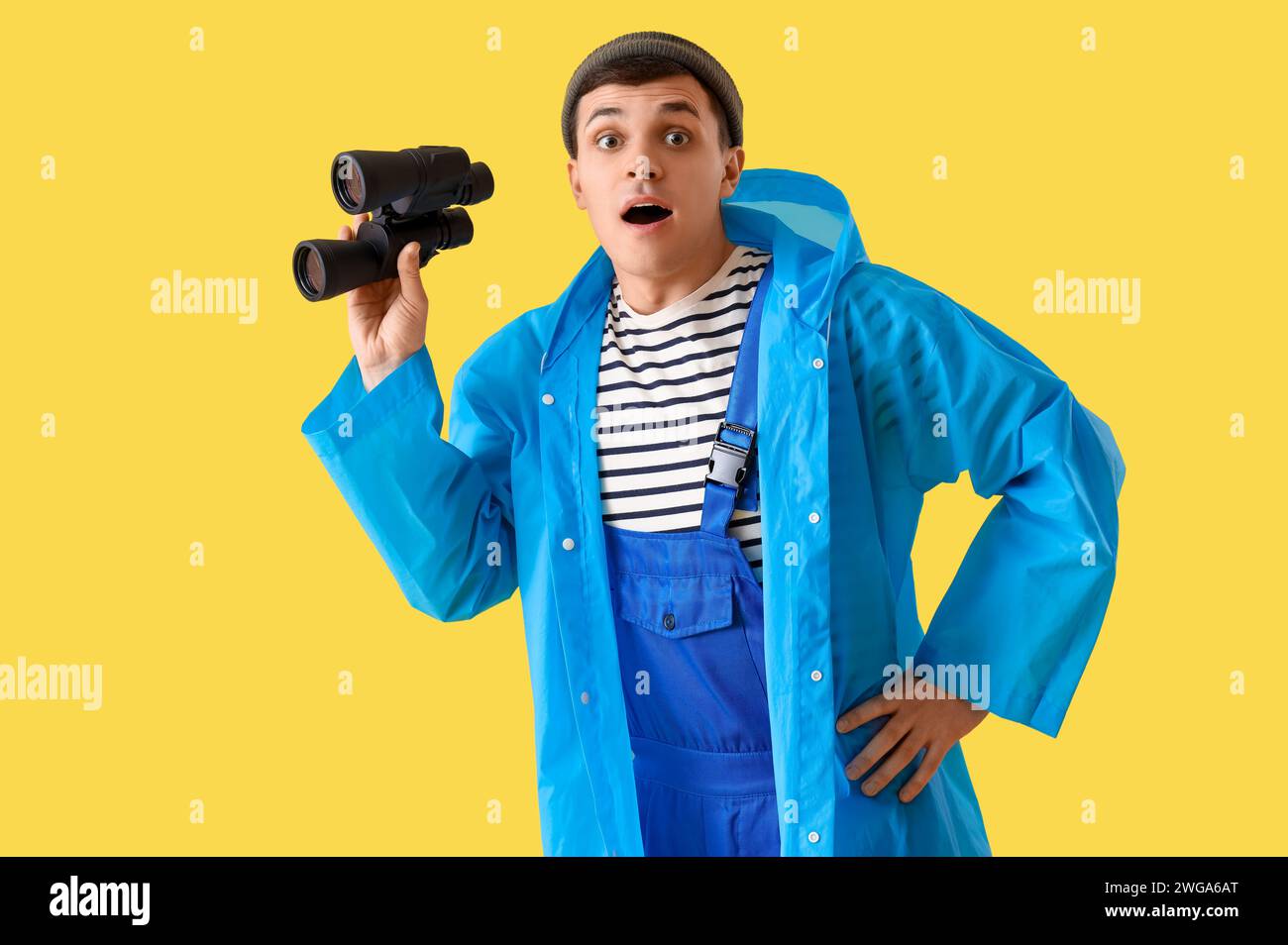 Surprised young sailor in raincoat with binoculars on yellow background ...