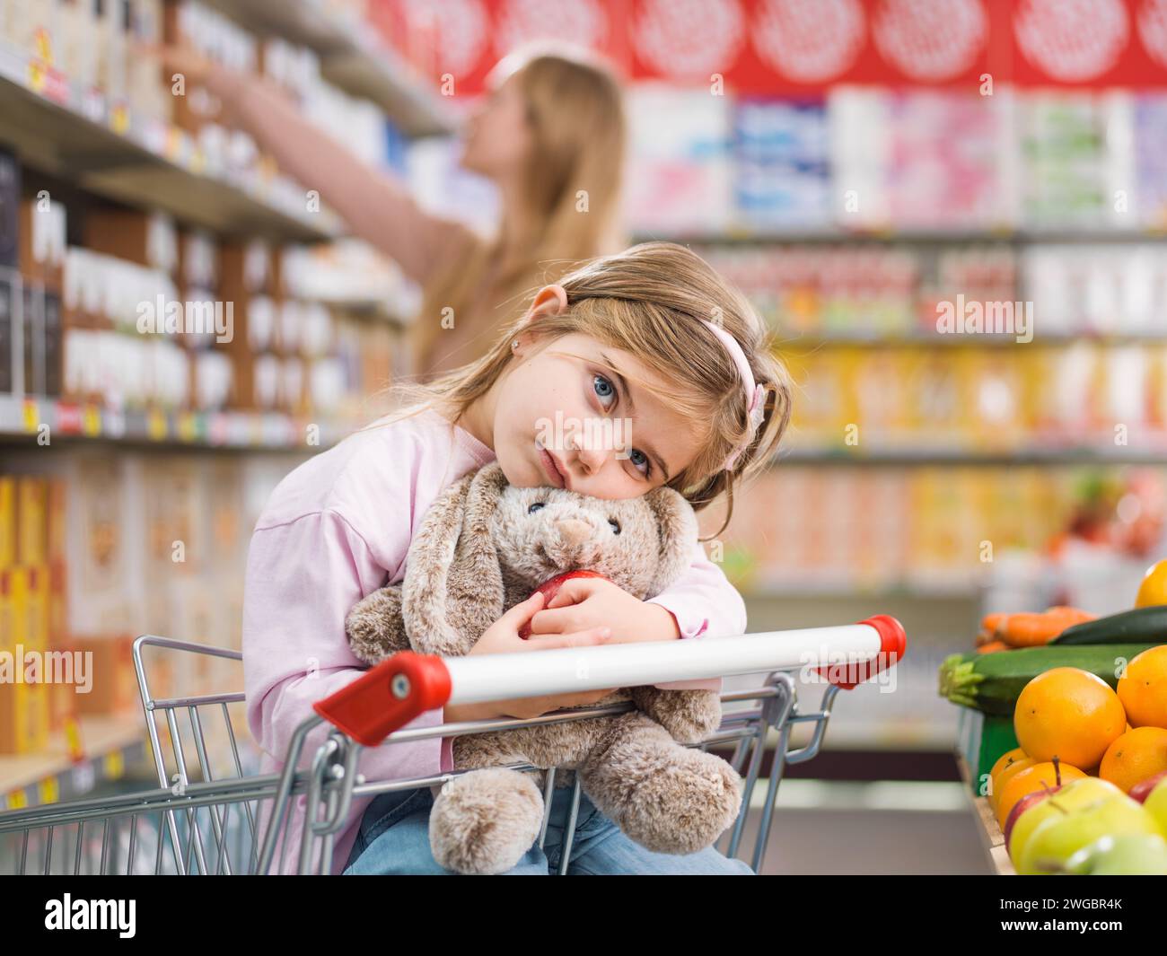 Cute bored child sitting on a shopping cart and hugging her plushie ...