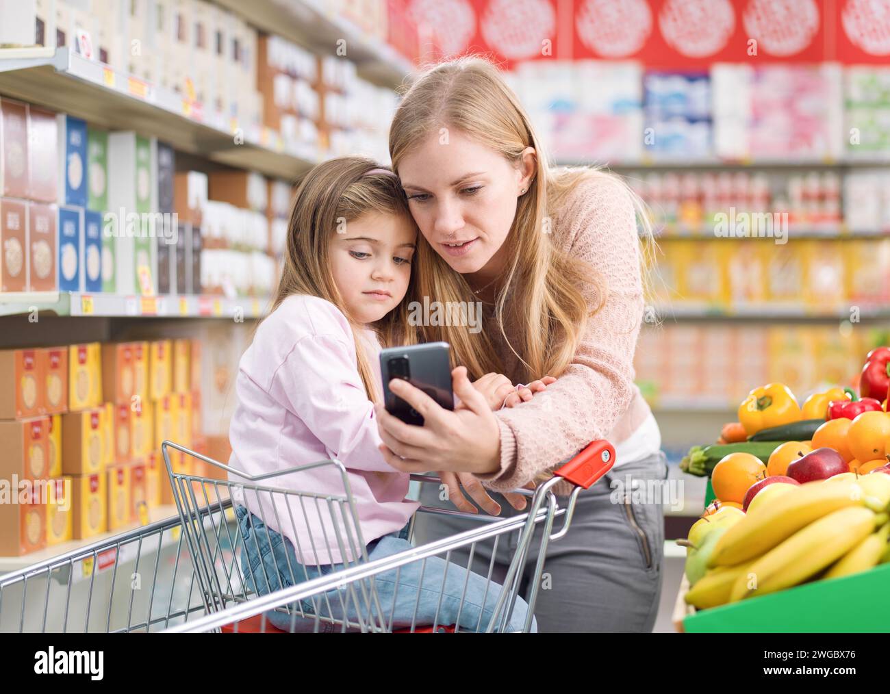 Cute girl sitting on a shopping cart at the supermarket, her mother is ...
