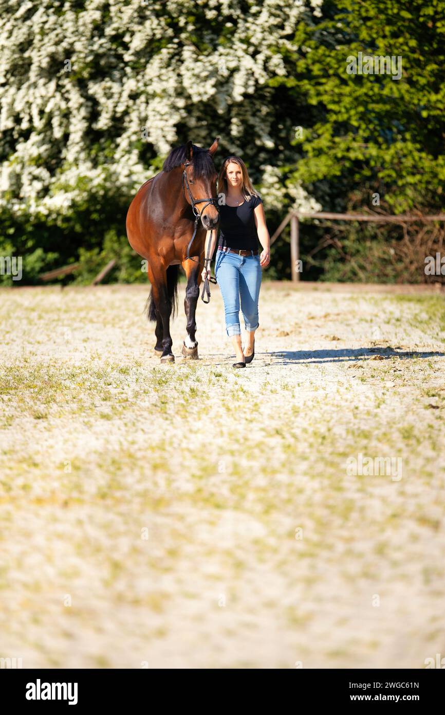 A young woman stands with her horse on a sunlit riding arena. In the background, blooming trees ...