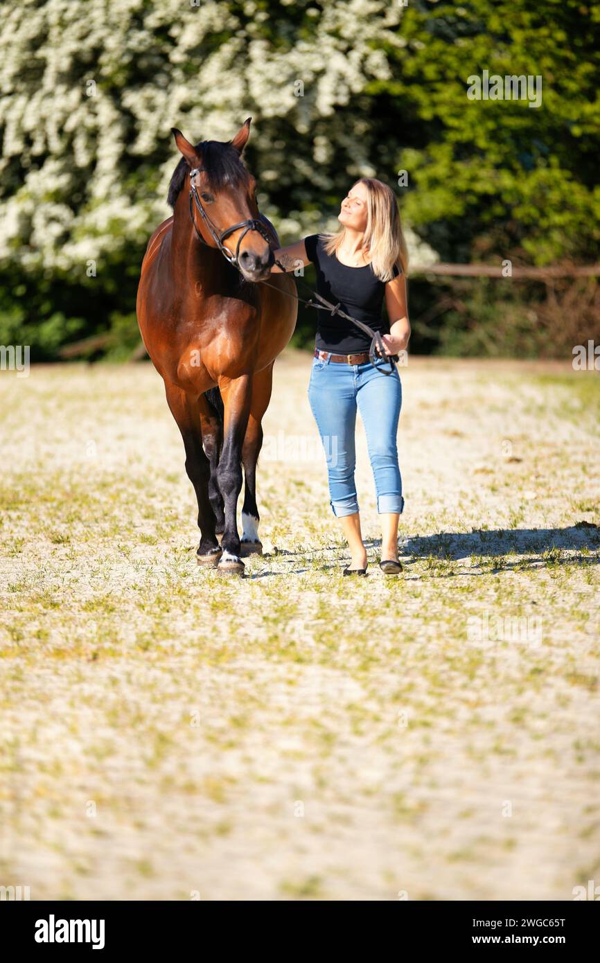 A young woman stands with her horse on a sunlit riding arena. In the background, blooming trees ...