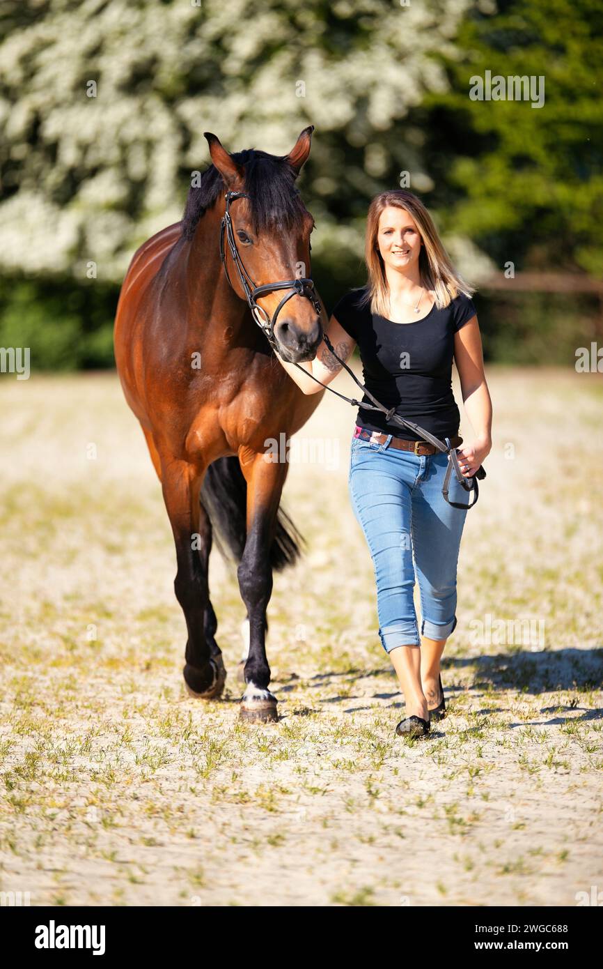 A young woman stands with her horse on a sunlit riding arena. In the background, blooming trees ...