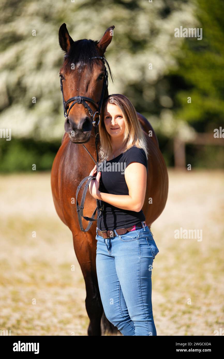 A young woman stands with her horse on a sunlit riding arena. In the background, blooming trees ...