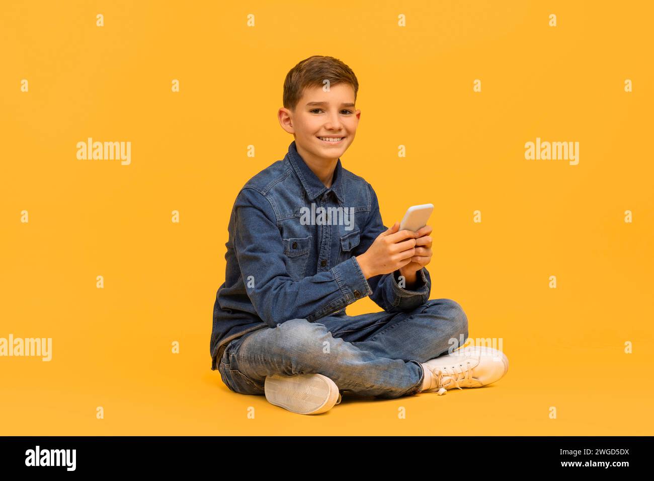 Smiling teen boy with smartphone in hands sitting cross-legged on yellow background Stock Photo ...