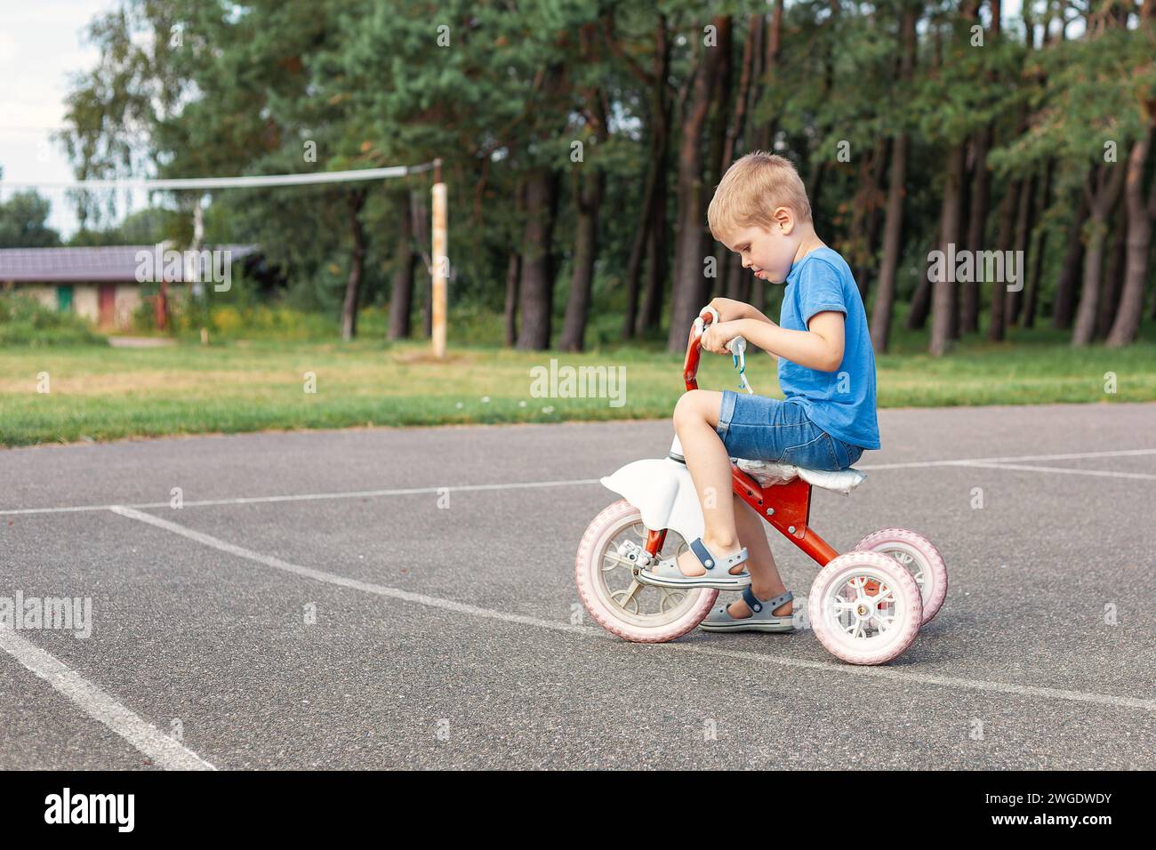 Very focused Little boy in blue, riding a red old tricycle in city park ...