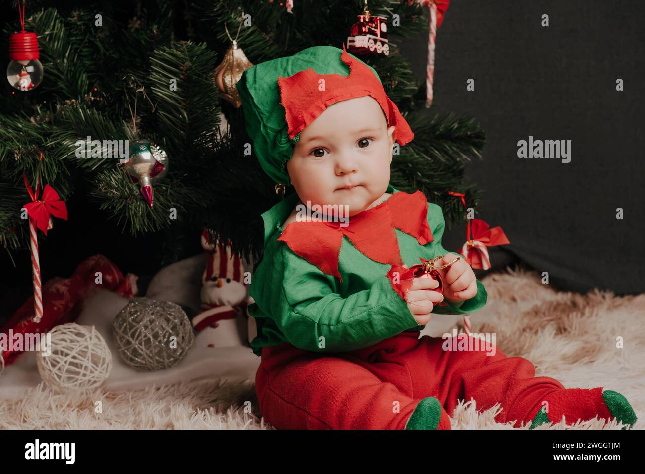 little boy in a red green elf costume sits under a Christmas tree with ...