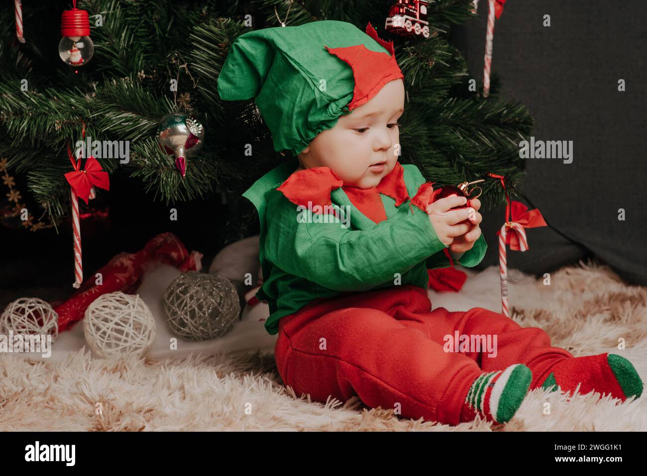 little boy in a red green elf costume sits under a Christmas tree with ...