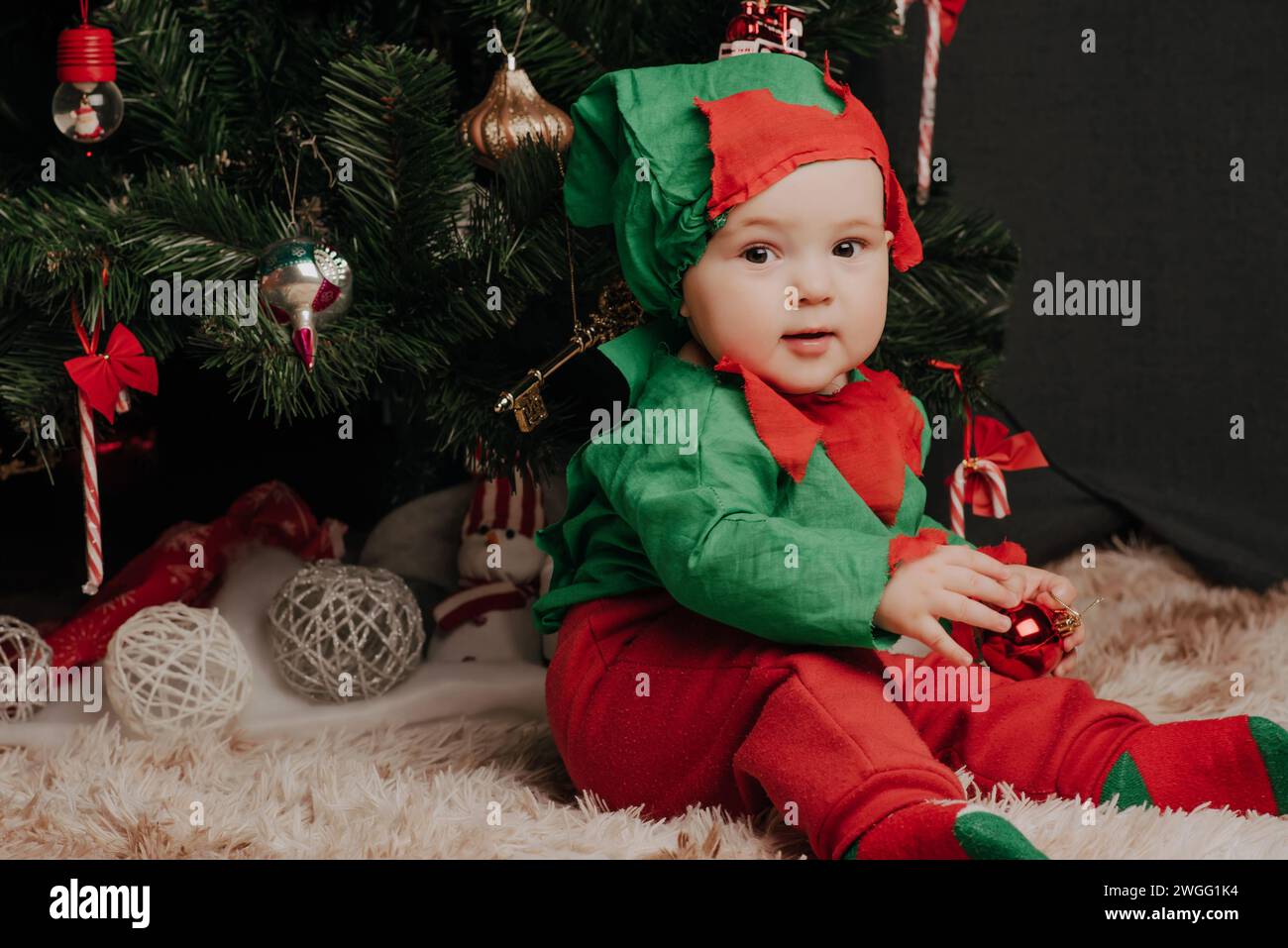 little boy in a red green elf costume sits under a Christmas tree with ...