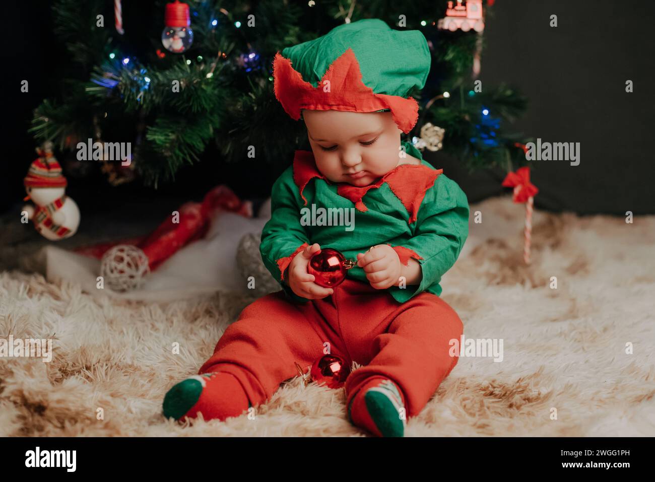 little boy in a red green elf costume sits under a Christmas tree with ...