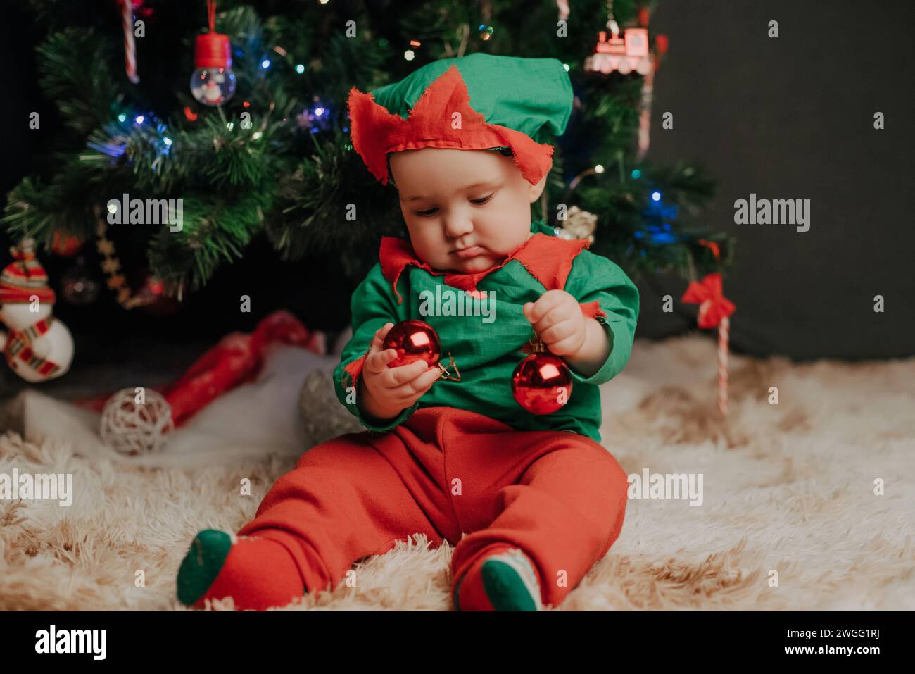 little boy in a red green elf costume sits under a Christmas tree with ...