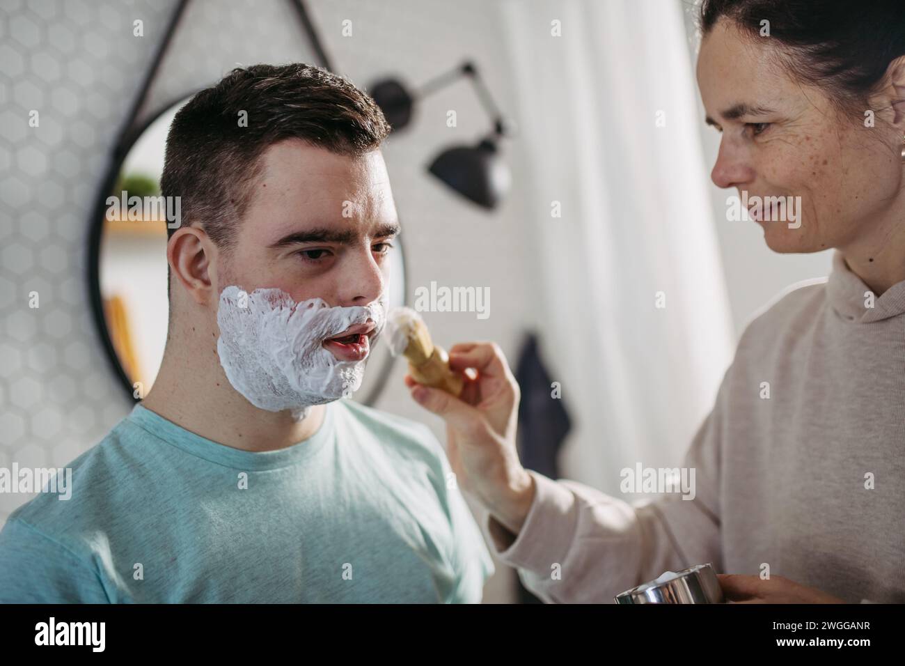 Mother applying shaving foam on son's face. Young man with down syndrome learning how to shave ...