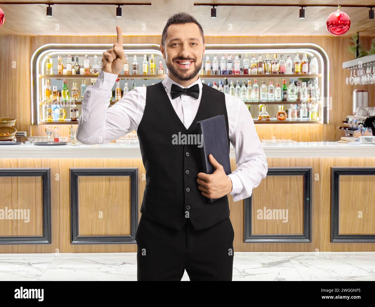 Cheerful waiter with a bow tie holding a menu and pointing up at a bar ...