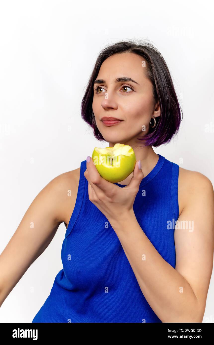 Positive woman holding bitten green apple. Looks to side. Blue dress ...