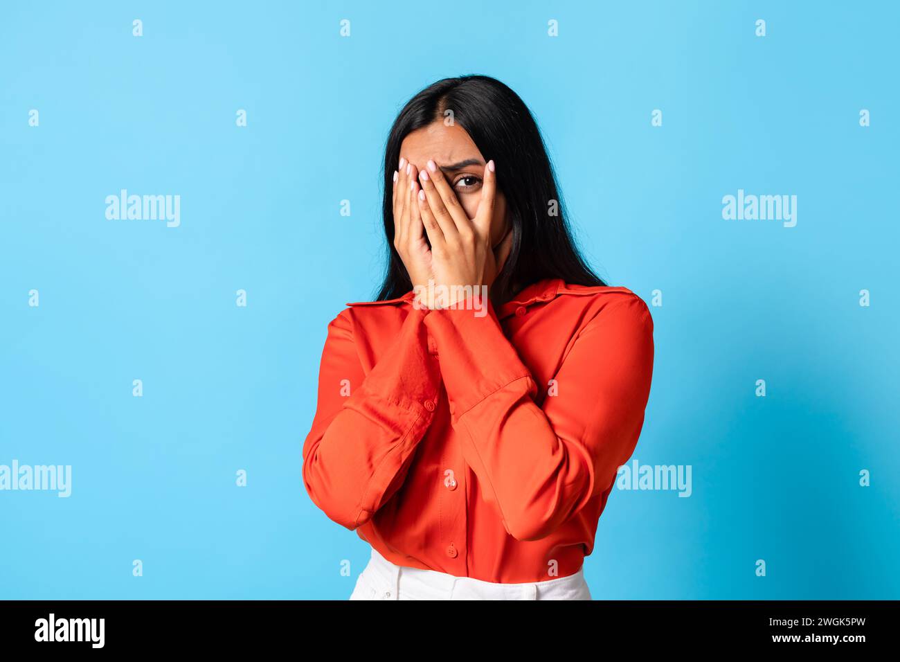 scared young indian woman peeking through fingers against blue backdrop ...