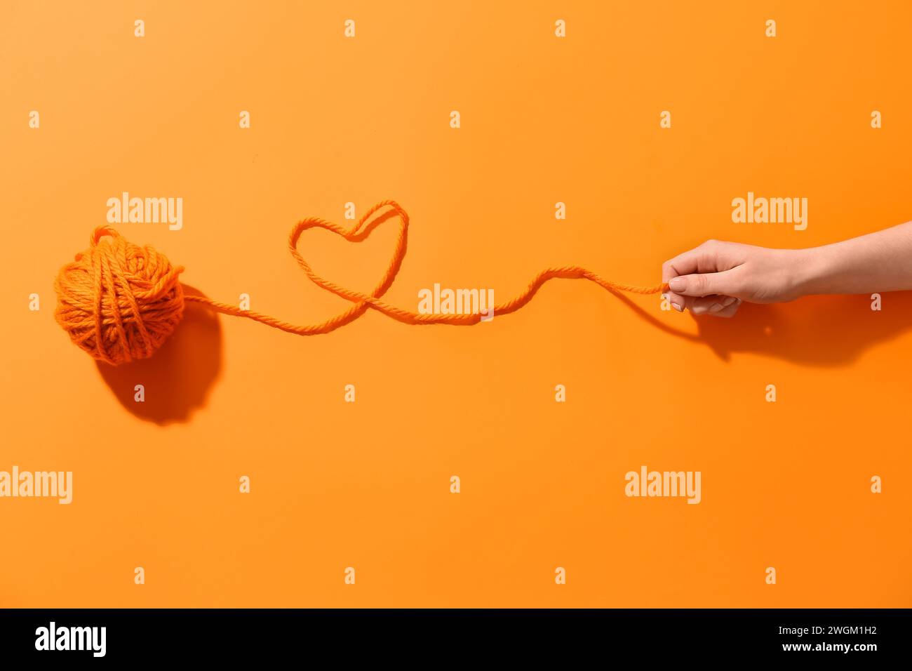 Female hand holding thread of yarn ball with heart on orange background ...
