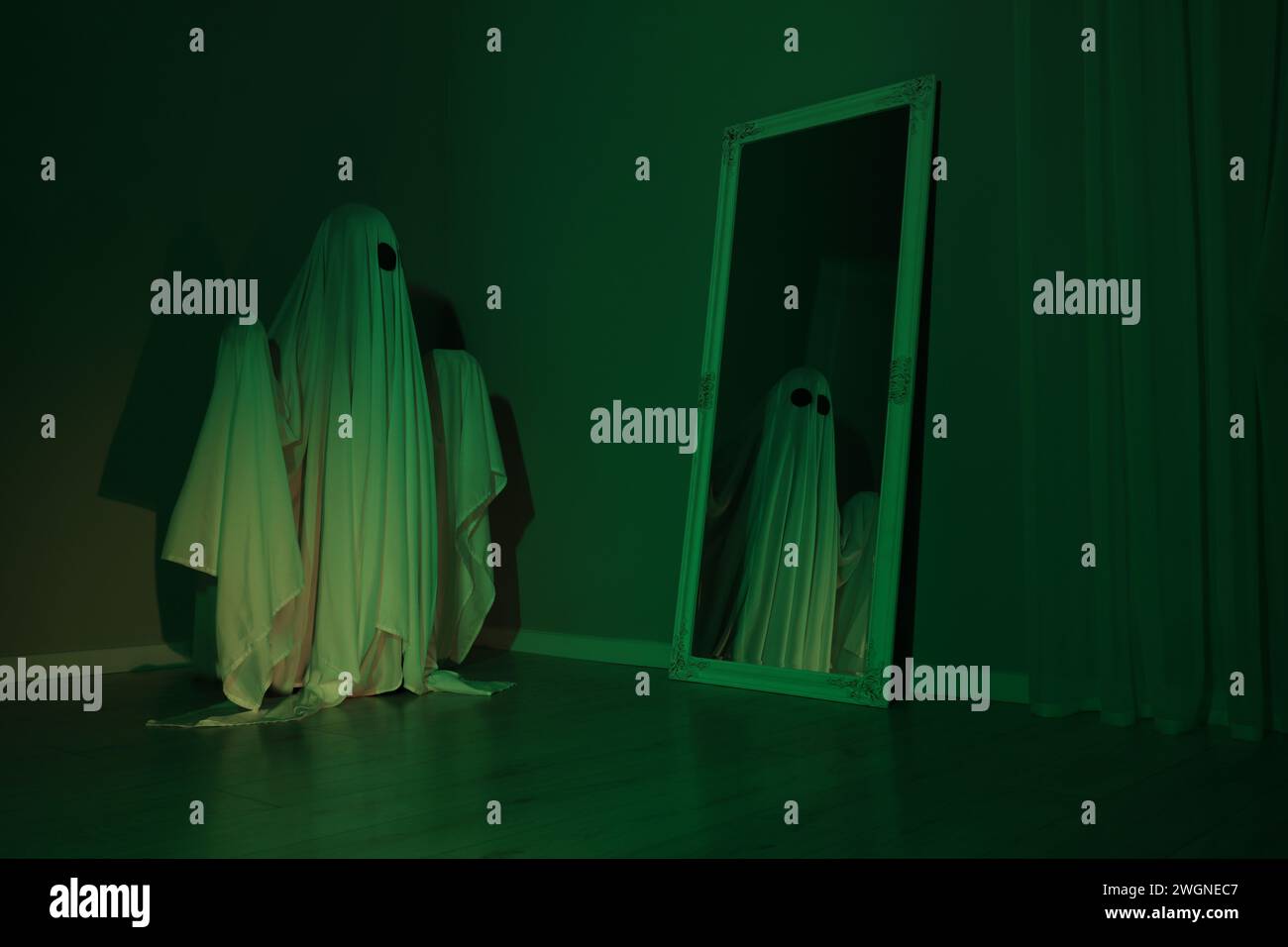 Creepy ghost. Woman covered with sheet looking at mirror in green light ...