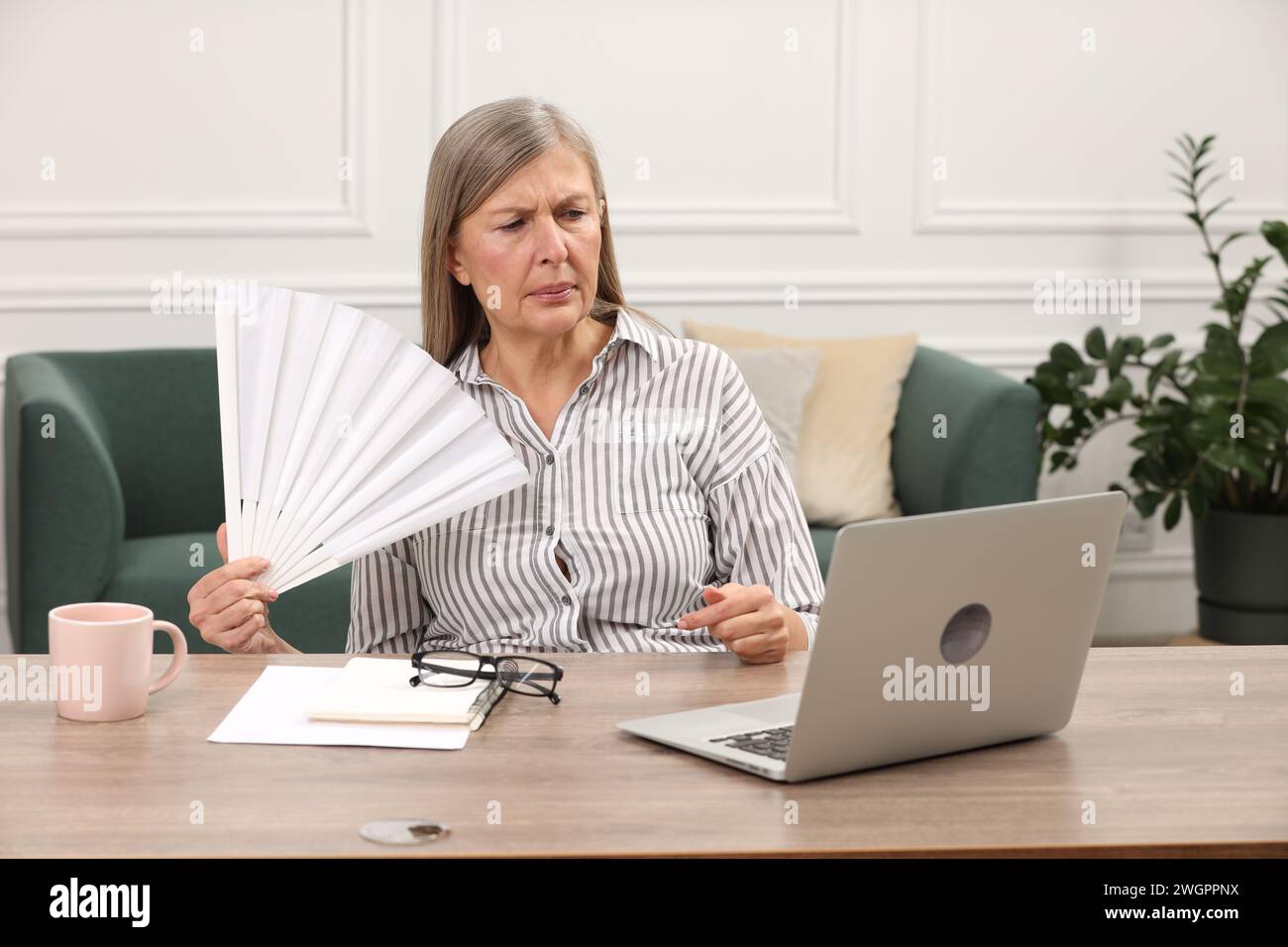 Menopause. Woman waving hand fan to cool herself during hot flash at ...