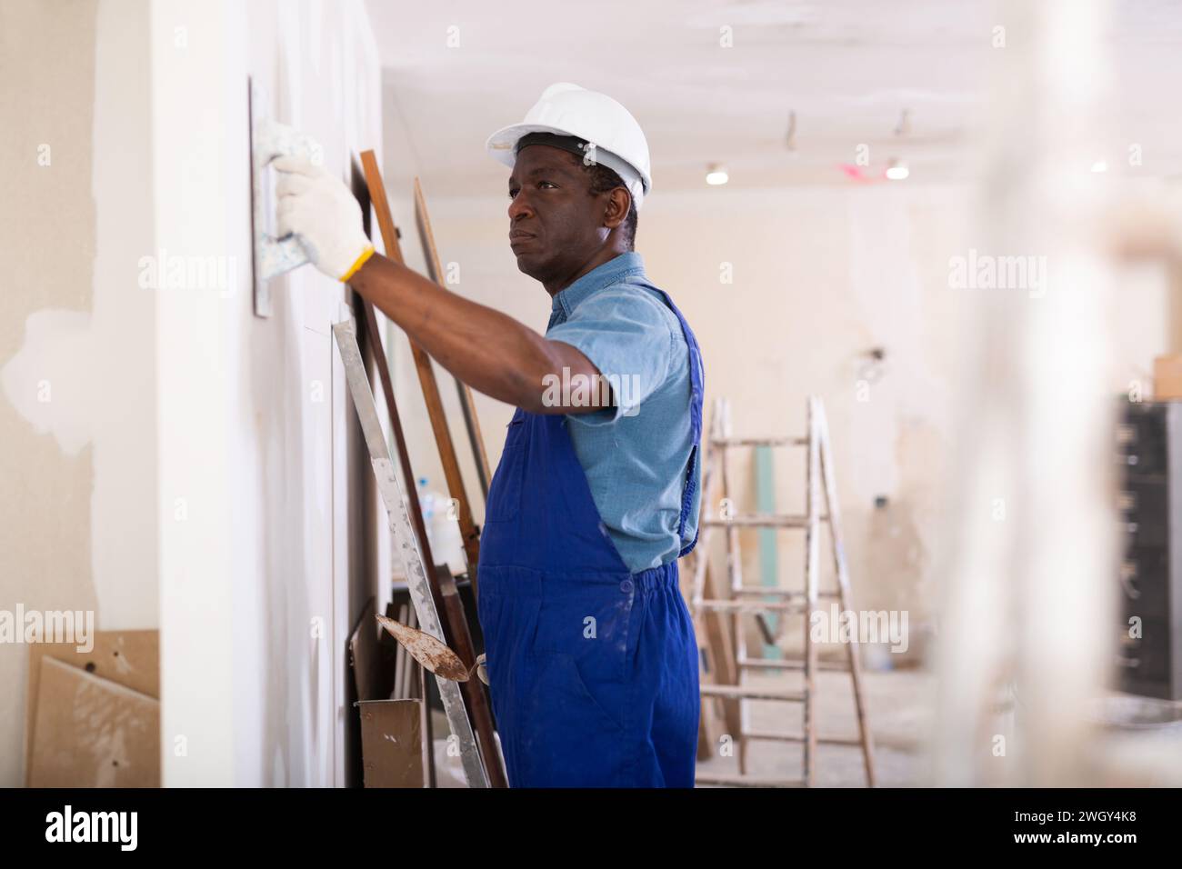 Painter in blue overalls leveling plaster on the wall with spatula ...