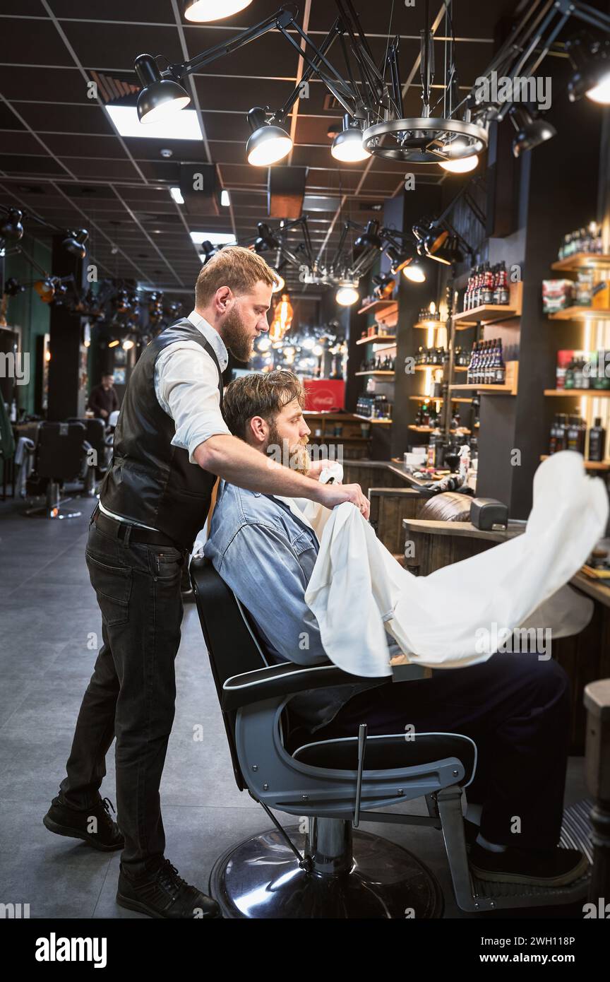 Fancy hairdresser putting on a cape on customer. Professional barber ...