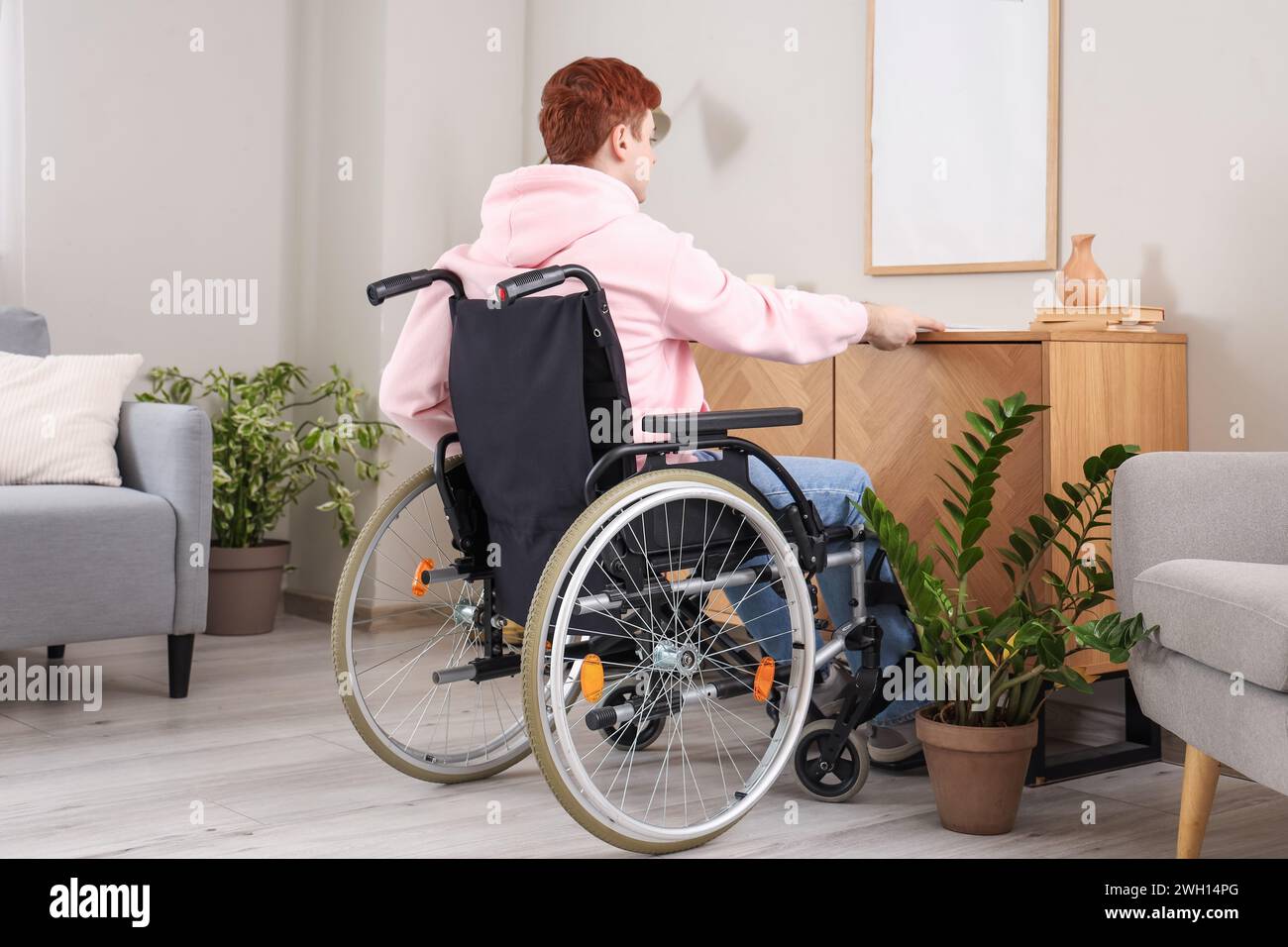 Young man in wheelchair taking magazine from shelf at home, back view ...