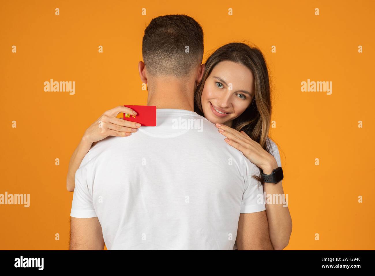 A smiling woman secretly holding a red credit card behind a man's back ...