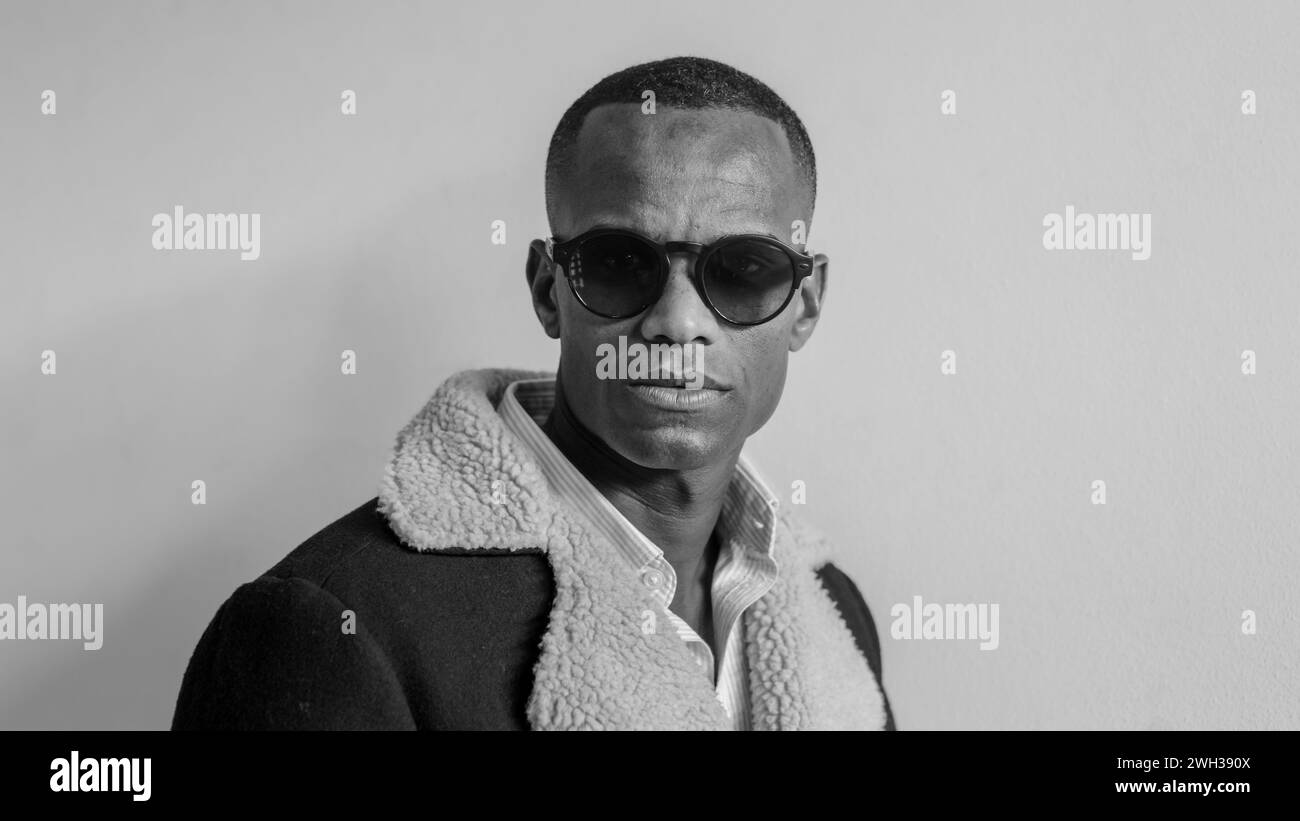 A man stands wearing sunglasses and a blue jacket, looking towards the ...