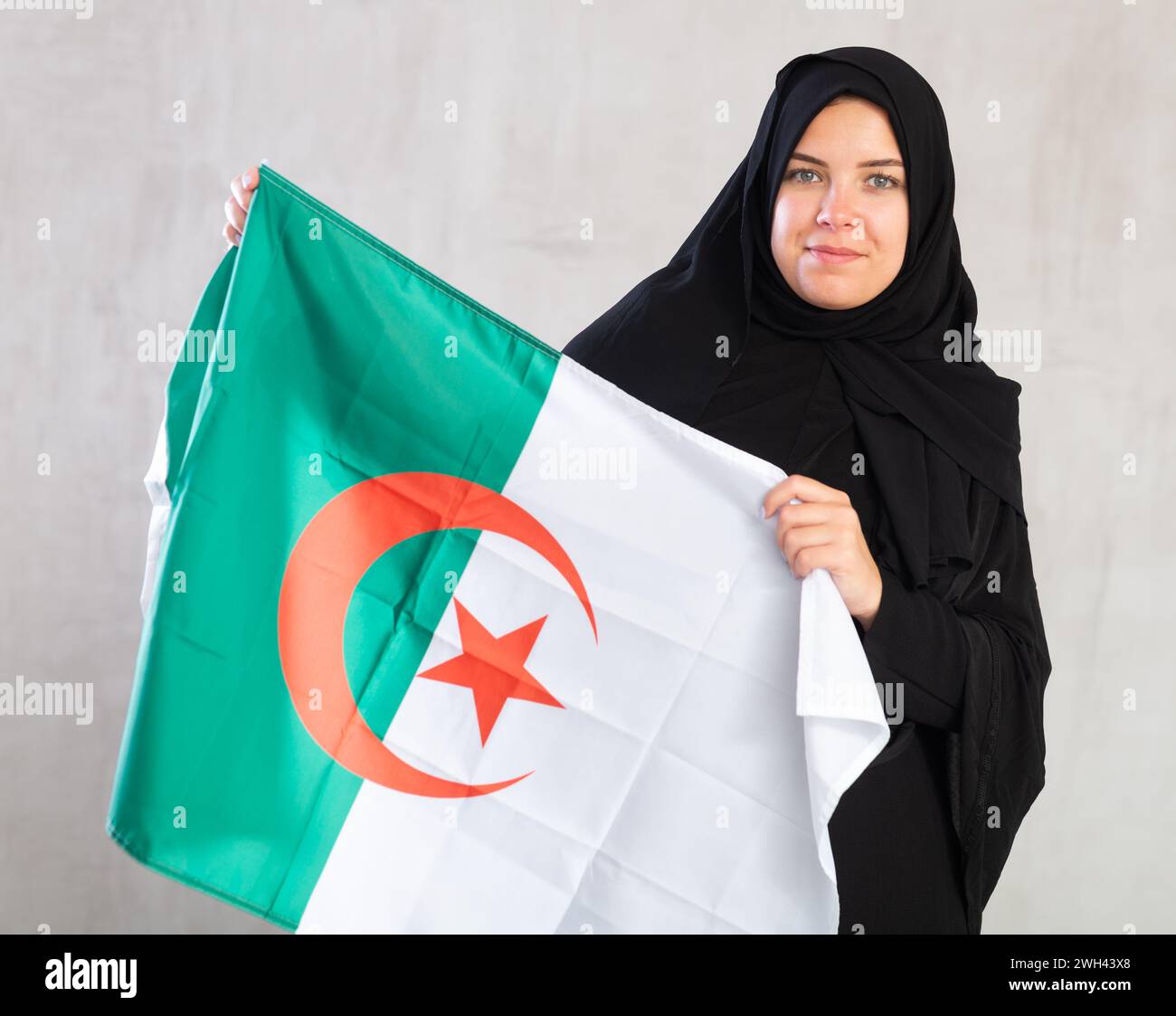 Balanced Muslim woman in traditional black hijab holds flag of Algeria ...