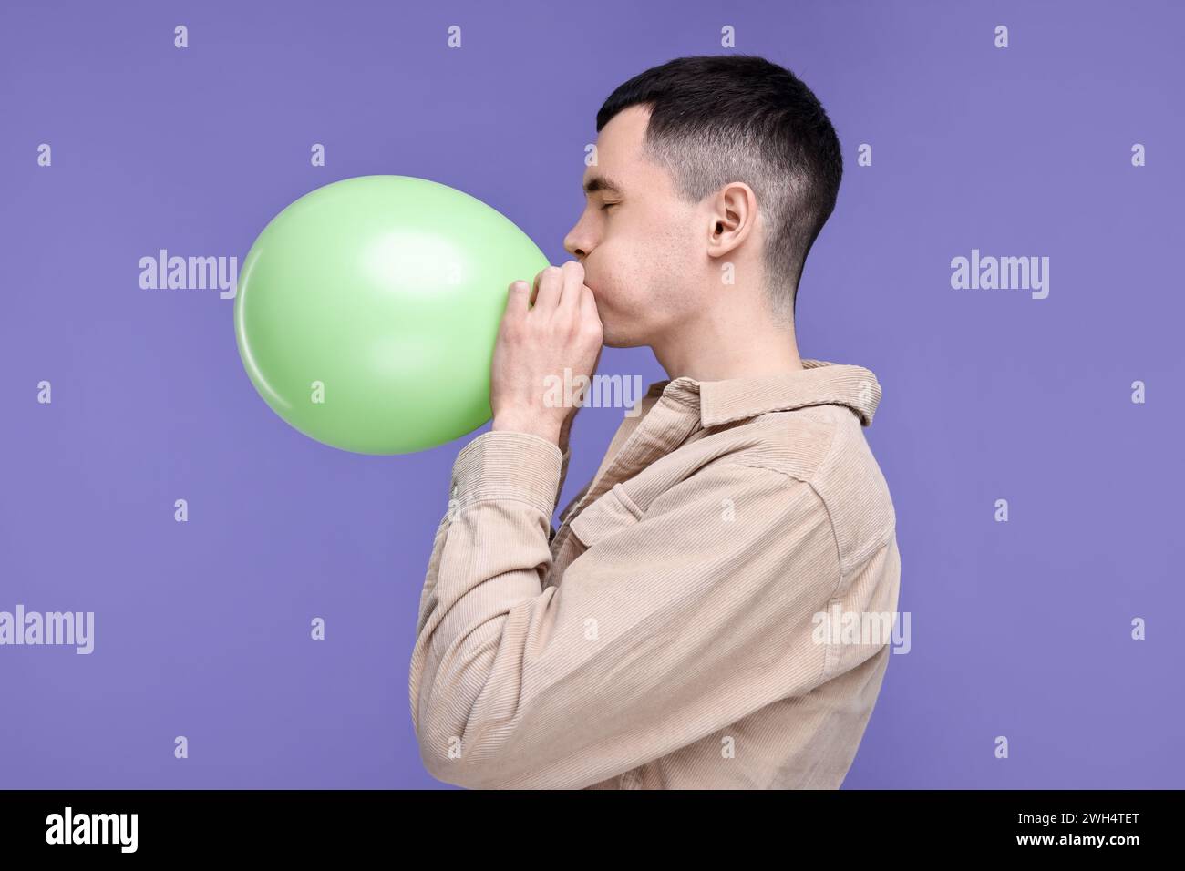 Young man inflating light green balloon on purple background Stock ...