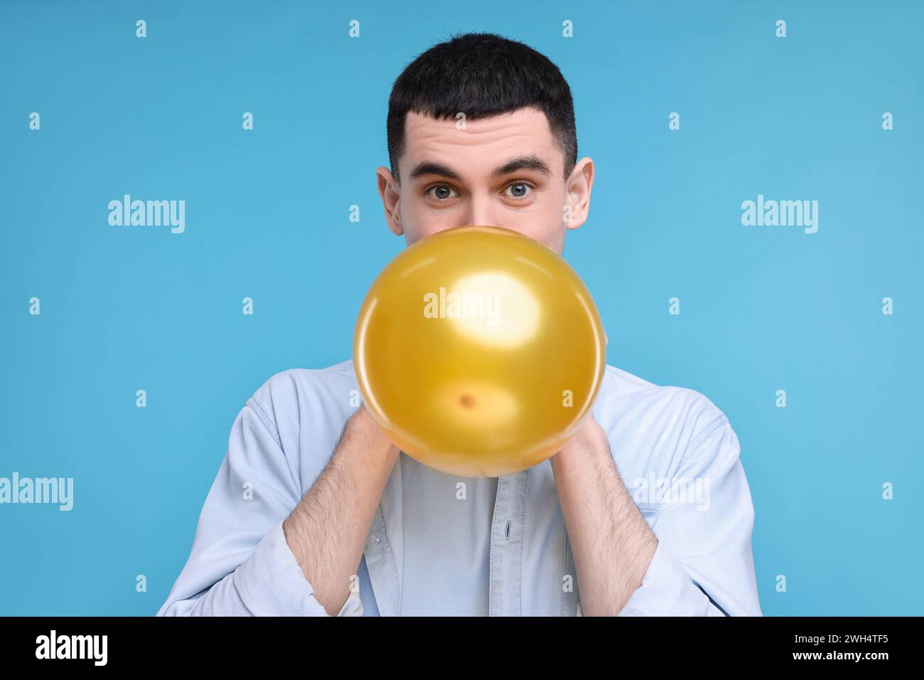 Young man inflating golden balloon on light blue background Stock Photo ...
