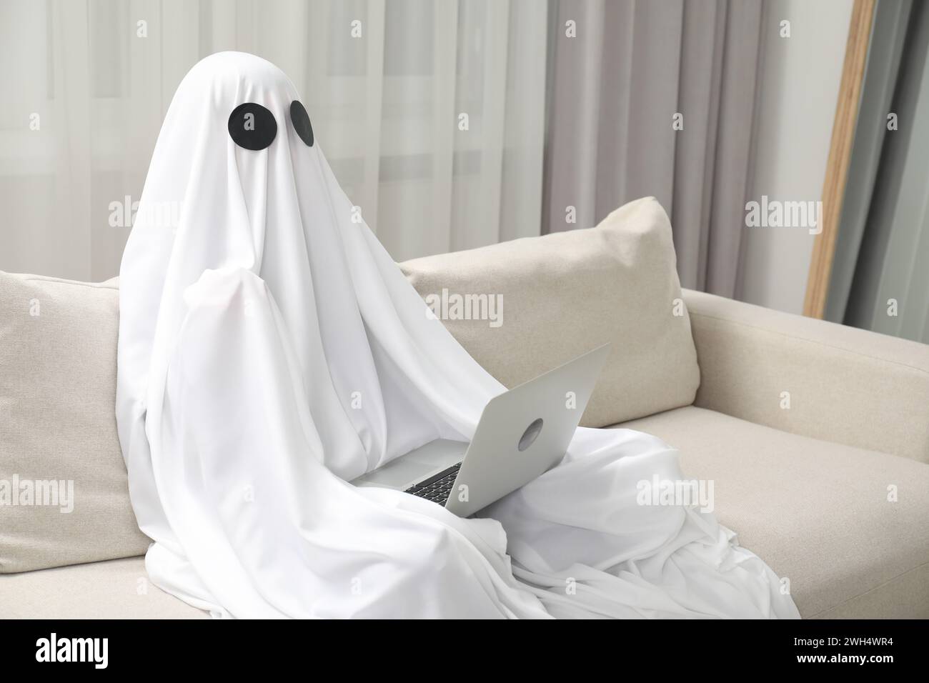 Creepy ghost. Person covered with white sheet using laptop on sofa at ...