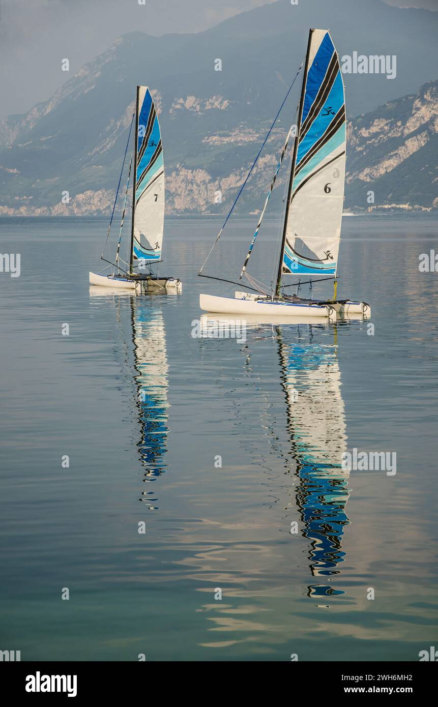 Small catamarans with blue and white sails reflected in Lake Garda ...