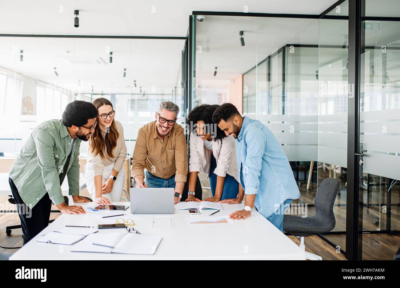 Diverse team huddles around table, and looks at a laptop screen in a bright office, closely ...