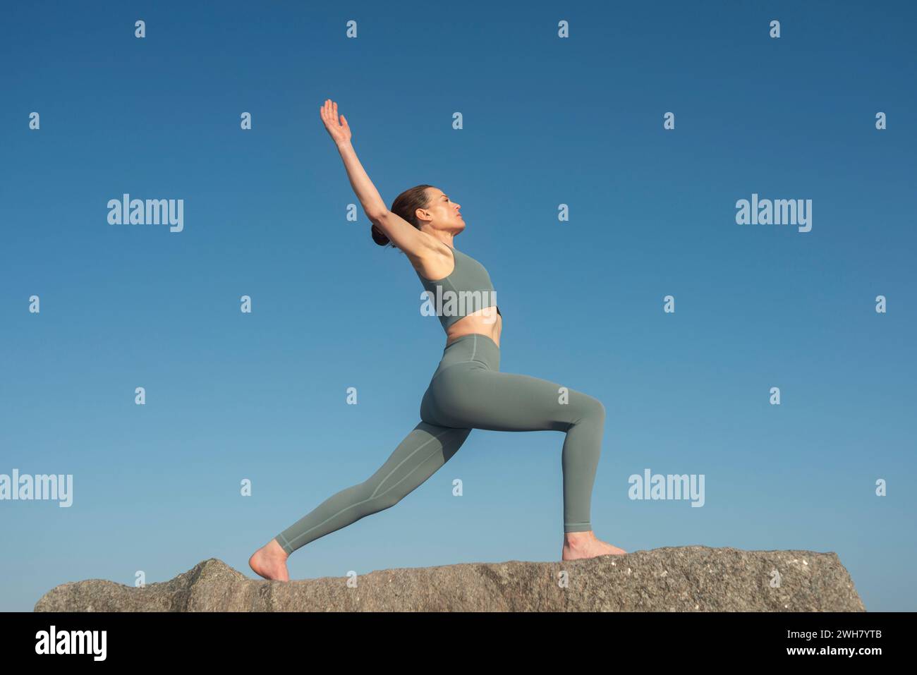 fit woman doing a warrior yoga pose standing on rocks, blue sky ...
