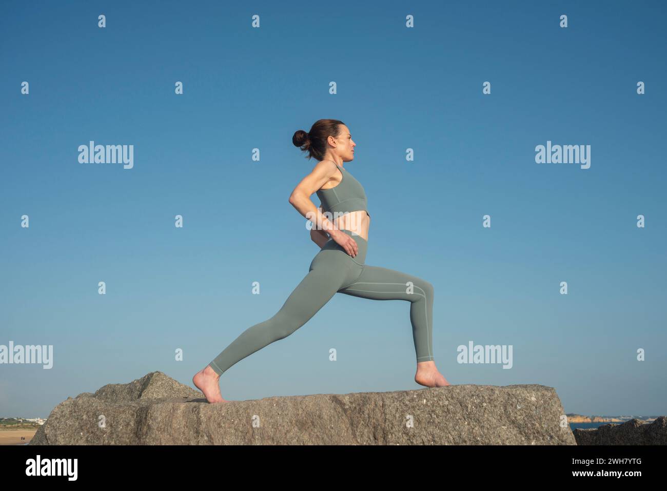 fit woman doing a high lunge yoga pose on rocks, blue sky background ...