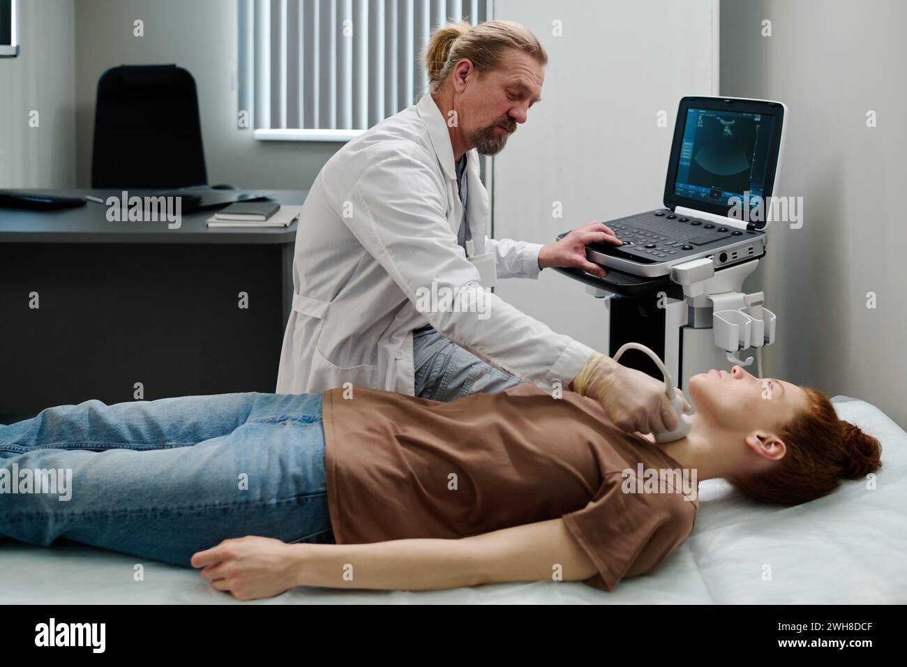 Mature endocrinologist in labcoat moving ultrasonic probe on neck of ...