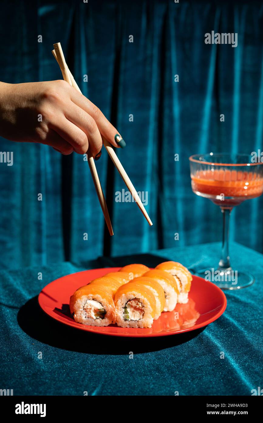 Female hand with wooden chopsticks taking sushi roll on a deep blue background. Asian restaurant ...