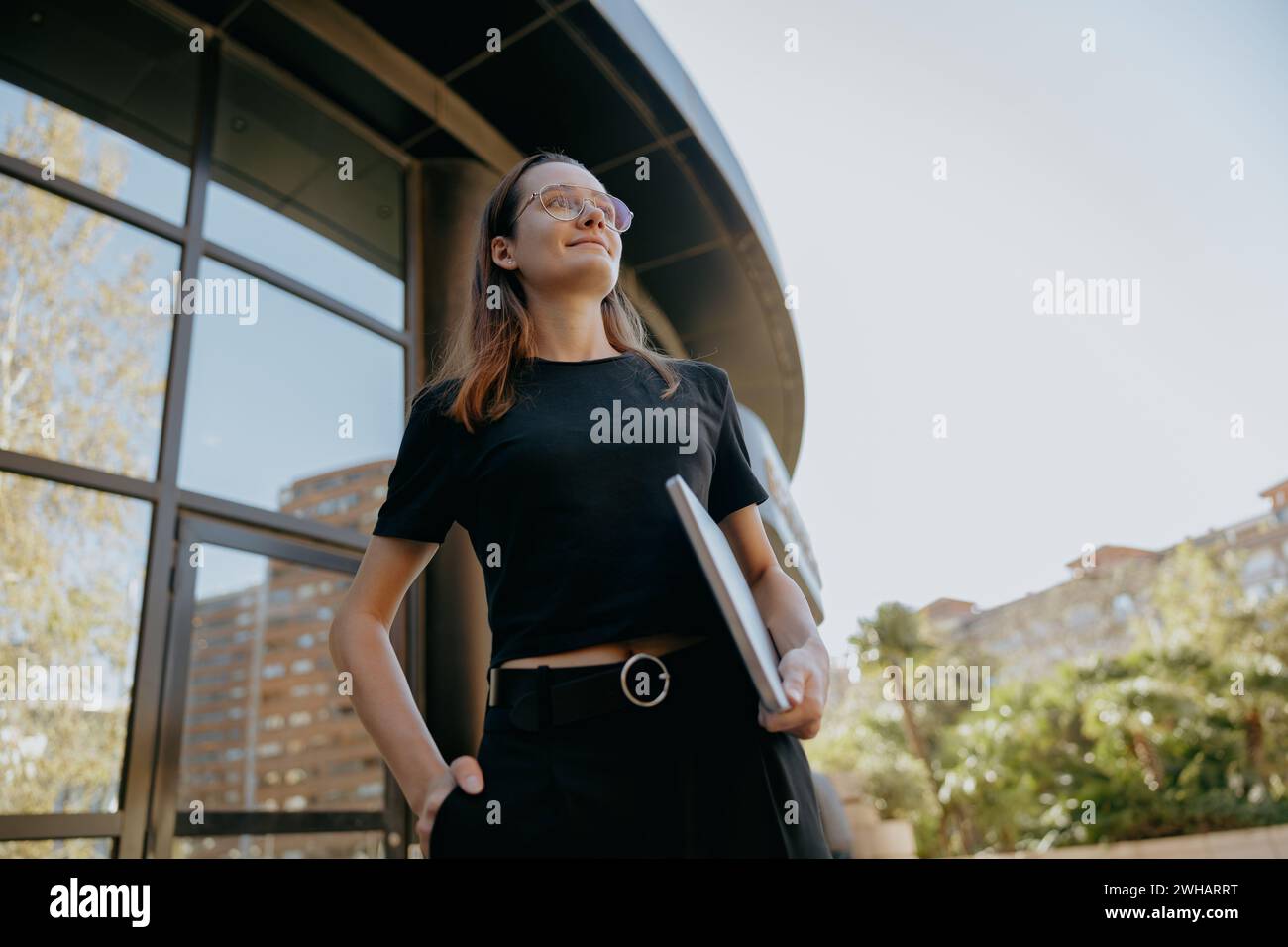 Young female freelancer with laptop standing on modern building ...