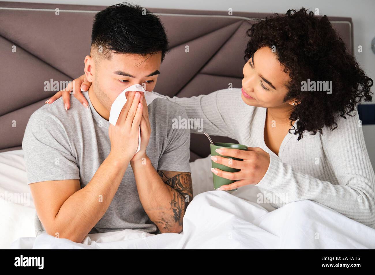Asian man blowing his nose in a tissue and his Latin wife taking care ...