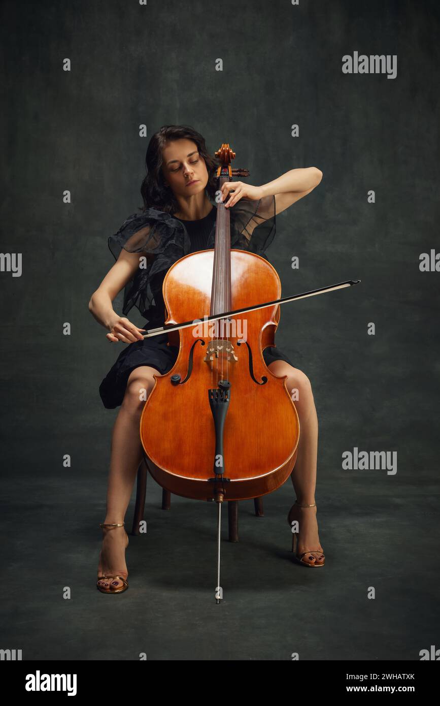 Tender young woman, cellist playing cello, sitting chair against dark ...