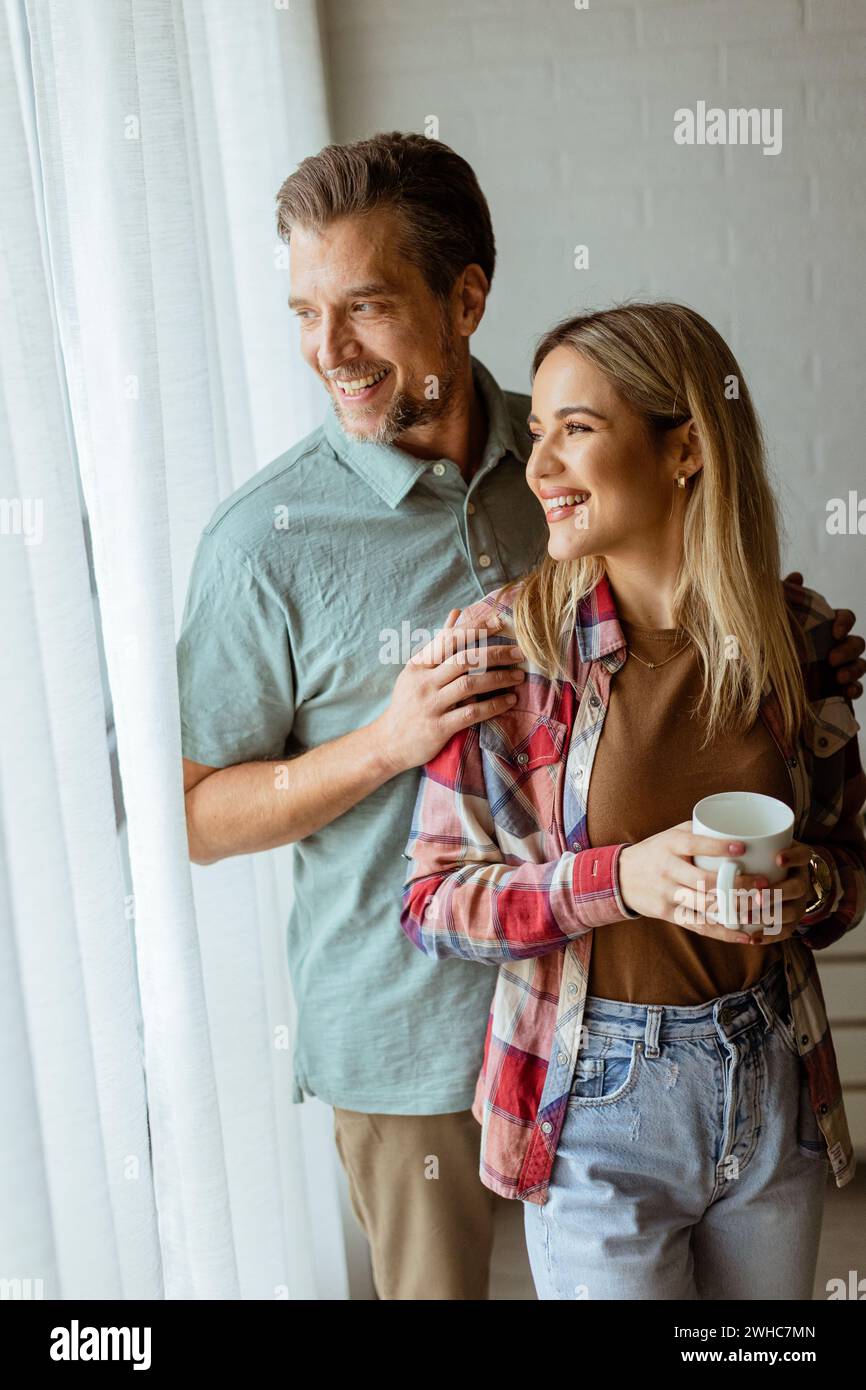 In a softly lit room, a man lovingly embraces a woman from behind as ...