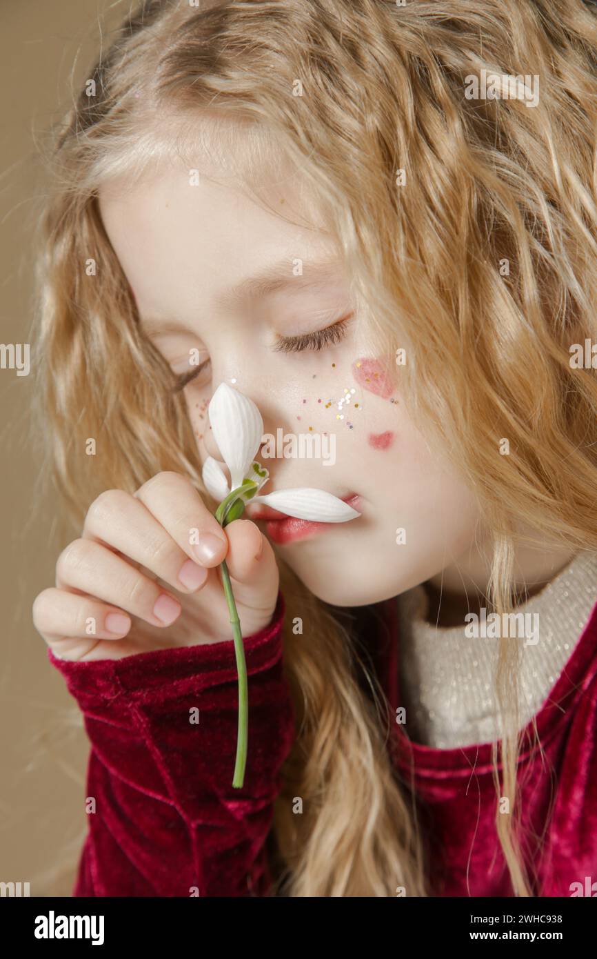 Portrait of cute little girl with glitters and red hearts on the face holding snowdrop flower ...