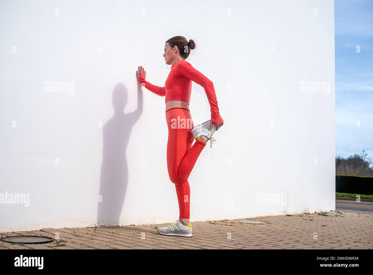 woman doing leg stretch exercise outdoors in the sun by a white wall ...
