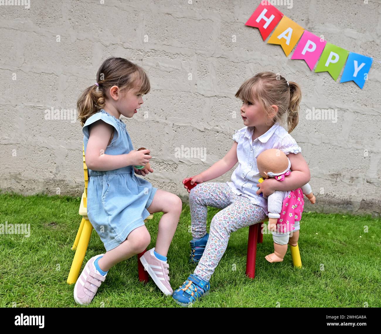 Two 3 year old girls sitting on chairs on grass in a garden talking ...