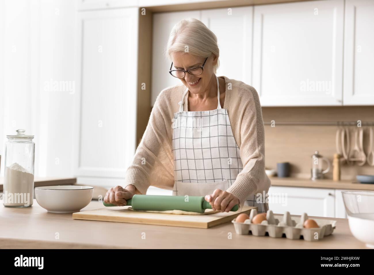 Positive mature baker woman wearing apron, rolling dough Stock Photo ...