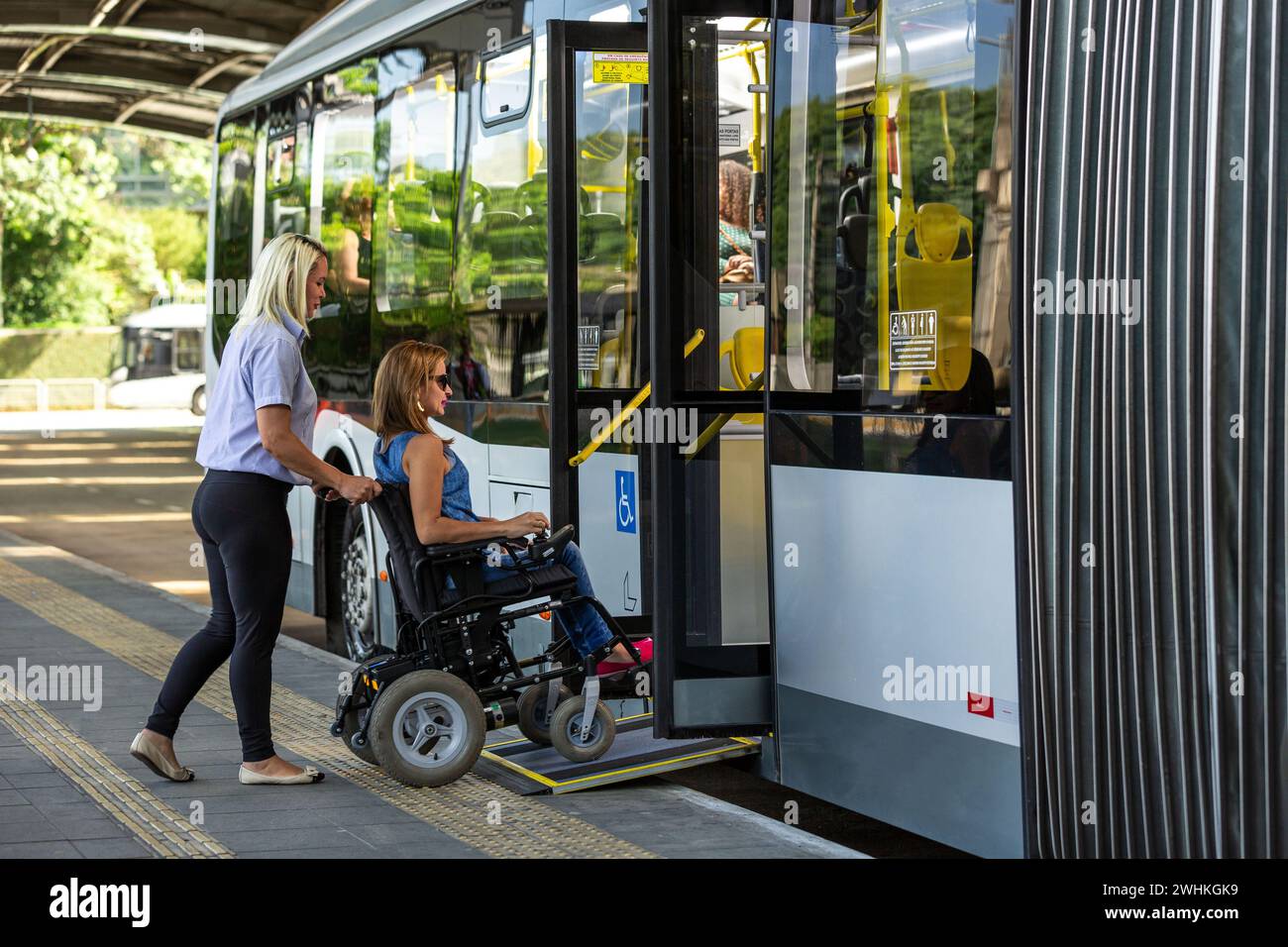Assistant assists wheelchair users to board a municipal bus via the access ramp, in a bi ...