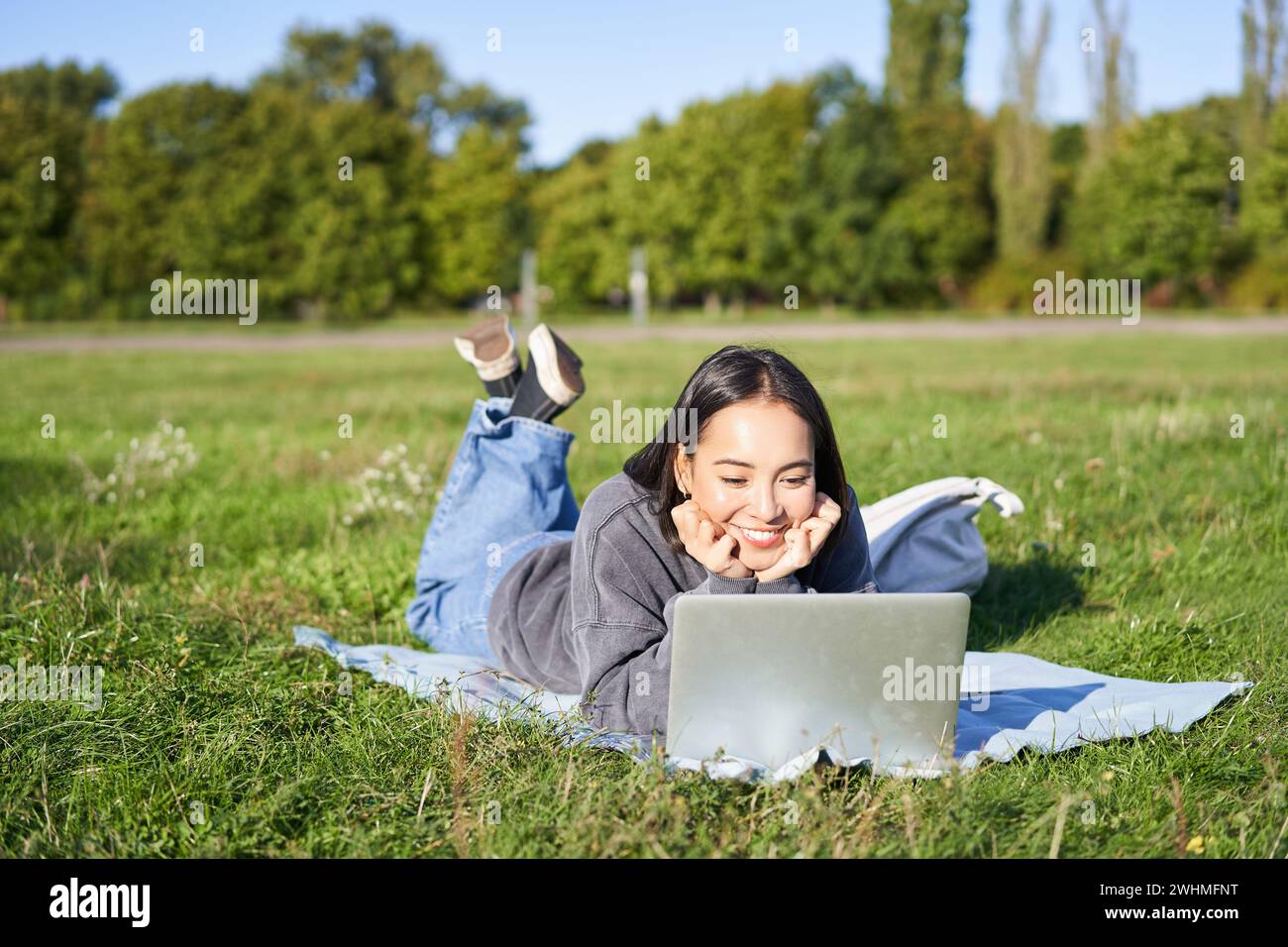 Smiling asian girl lying in park on grass, watching movie or video on ...