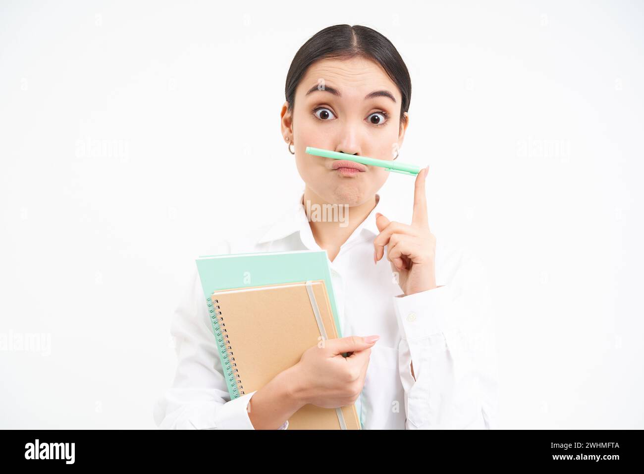 Funny korean woman with notebooks, holds pen with lips and nose and makes faces, stands over ...